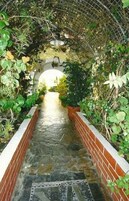 Uma passagem de pedra serpenteia por um túnel verdejante e exuberante, com paredes de tijolo e um arco de metal.