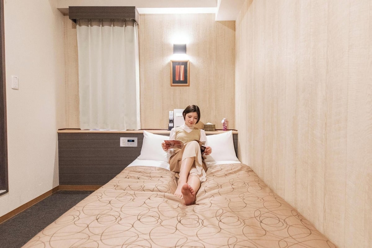 Quarto de hotel estreito com cama, mesa de cabeceira, espelho e luminária fixada na parede.