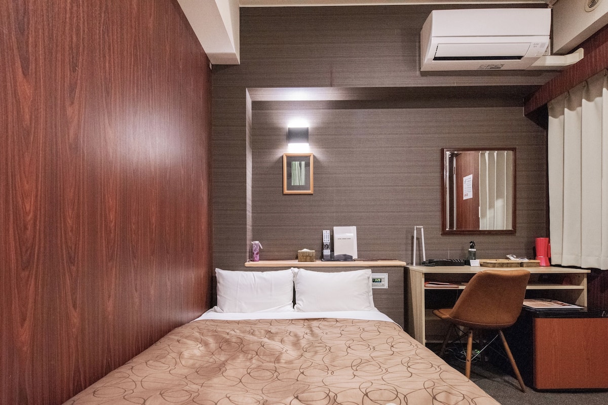 Quarto de hotel com cama, escrivaninha, cadeira, espelho e ar condicionado.
