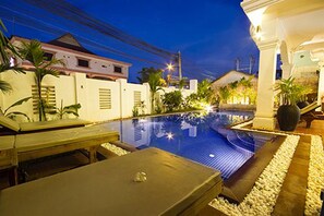 Uma área de piscina à noite, com piscina transparente, rodeada por paredes brancas e palmeiras, e um edifício ao fundo.