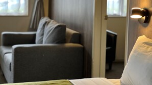Quarto de hotel com cama, sofá cinza e luminária fixada na parede.