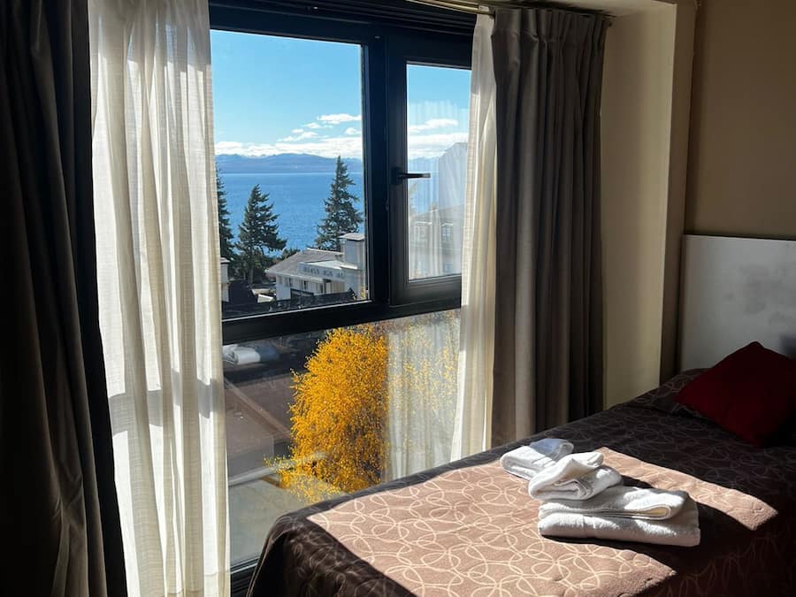 Quarto de hotel com cama, janela com cortinas e vista para o mar e montanhas.