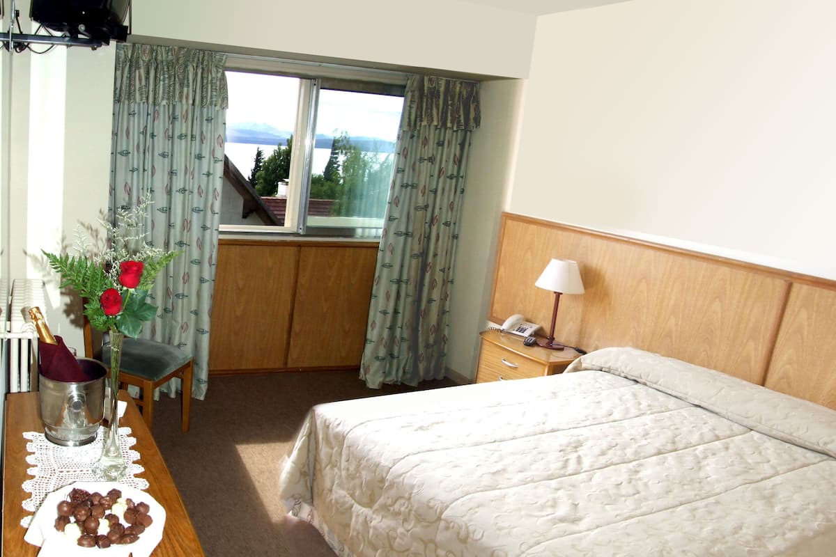 Quarto de hotel com cama, escrivaninha com uma garrafa de champanhe, vaso com flores, cadeira, mesa de cabeceira com abajur e janela com cortinas.