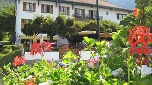 Um edifício branco com telhado em tijolos, rodeado por vegetação exuberante e flores vermelhas.
