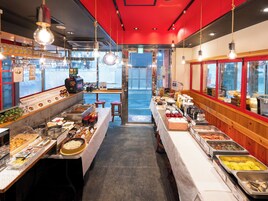 Buffet montado com diversas estações de comida, uma máquina de café e área de estar com bancos.