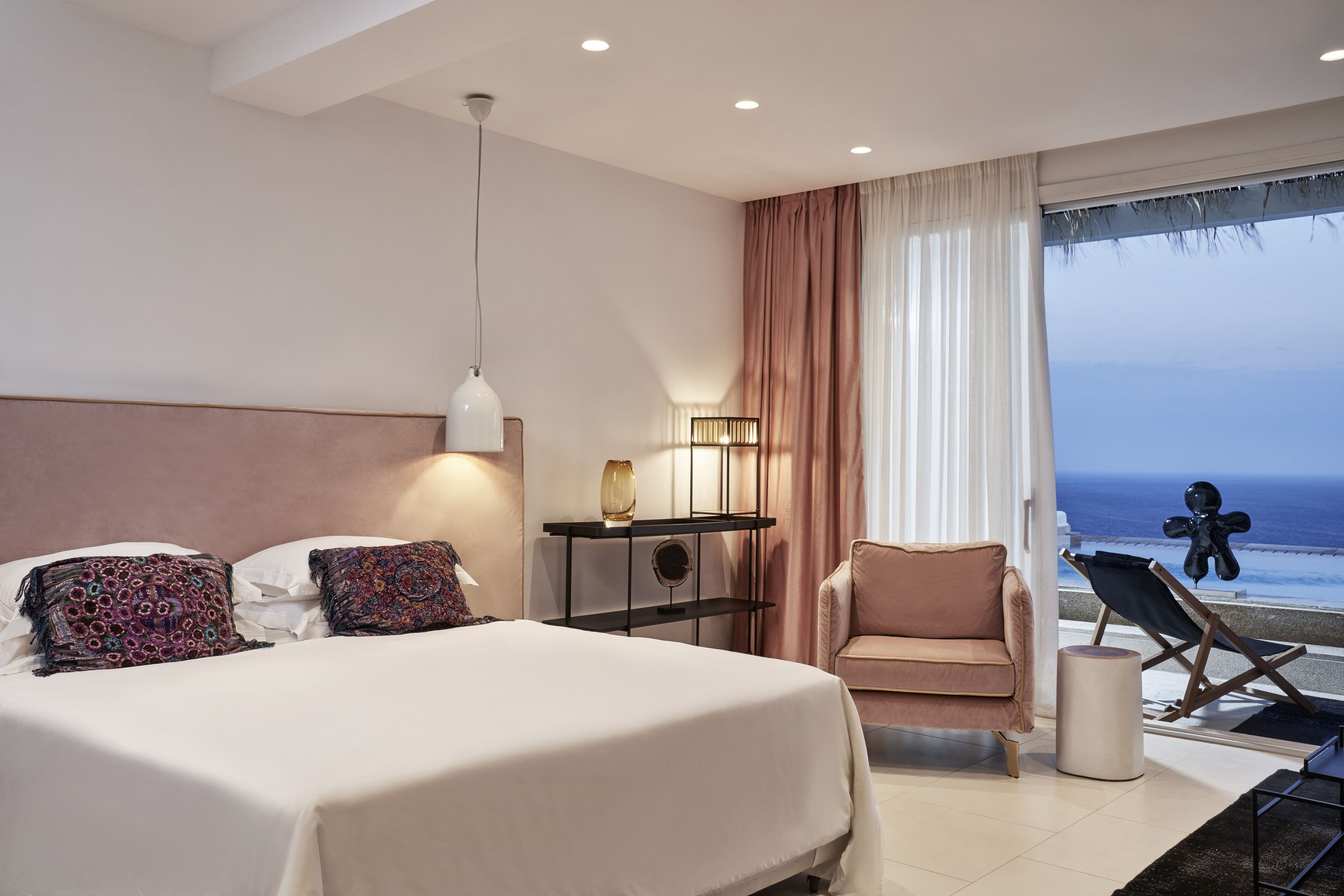 Quarto de hotel com uma cama, uma cadeira, uma mesinha e vista para o oceano.