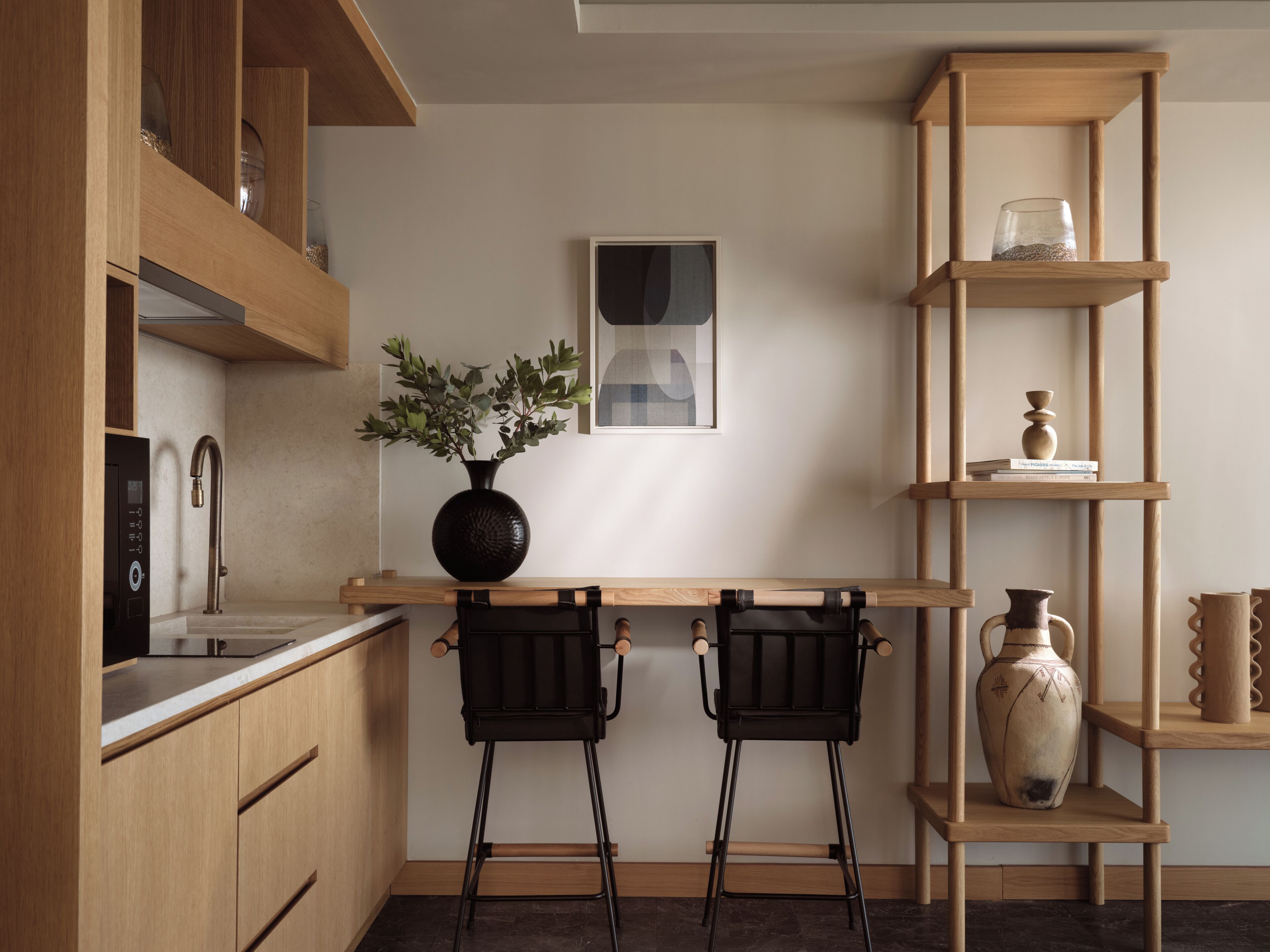 Cozinha moderna com armários de madeira, um vaso preto com plantas verdes, duas cadeiras pretas e uma prateleira de madeira com objetos decorativos.