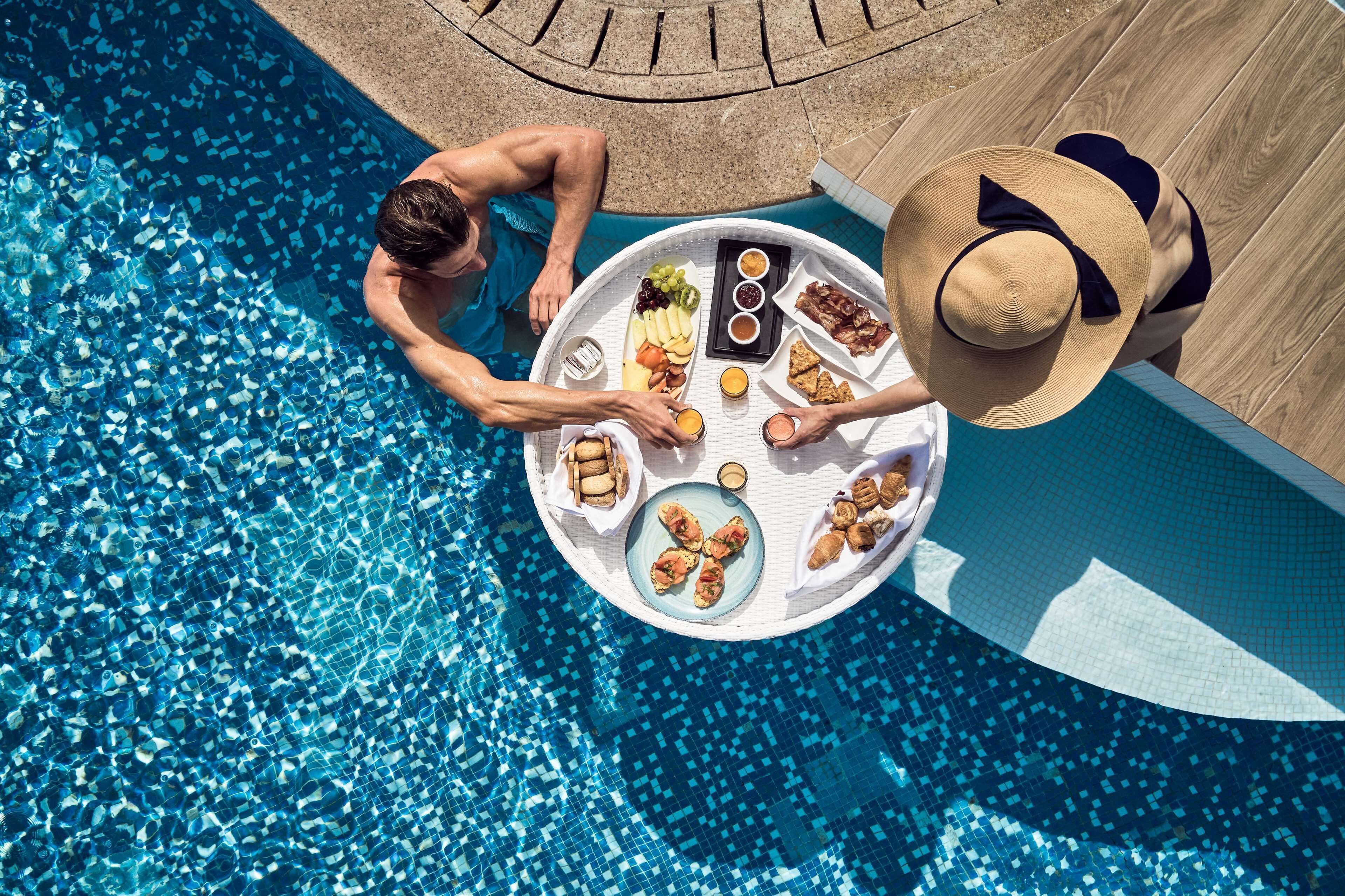 Vista aérea de uma área de refeições à beira da piscina, com uma mesa posta para dois, em volta de uma piscina azul cristalina.