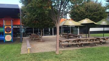 Parque com parquinho infantil, incluindo escorregas, estrutura de escalada, mesas para piquenique e guarda-sóis identificados com a inscrição "Cascade".