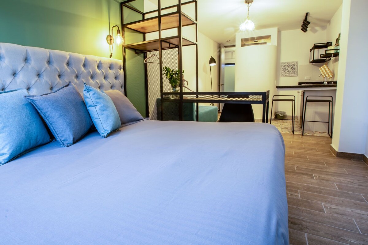 Quarto com cabeceira estofada, cama com roupa de cama azul, escrivaninha com cadeira e planta pendente.