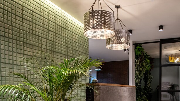 Uma área de recepção com parede revestida de azulejos verdes, balcão de atendimento, uma grande planta em vaso e luminárias pendentes.