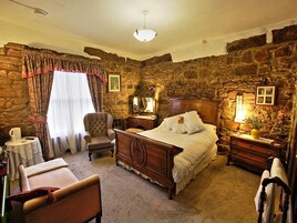 Quarto com parede de pedra, cama de madeira, uma cadeira, mesa de cabeceira e abajur.