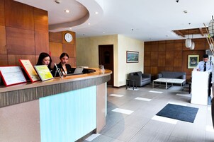 Uma área de recepção de hotel com balcão, dois recepcionistas, um relógio, um quadro emoldurado e uma área de estar com sofá e mesa.