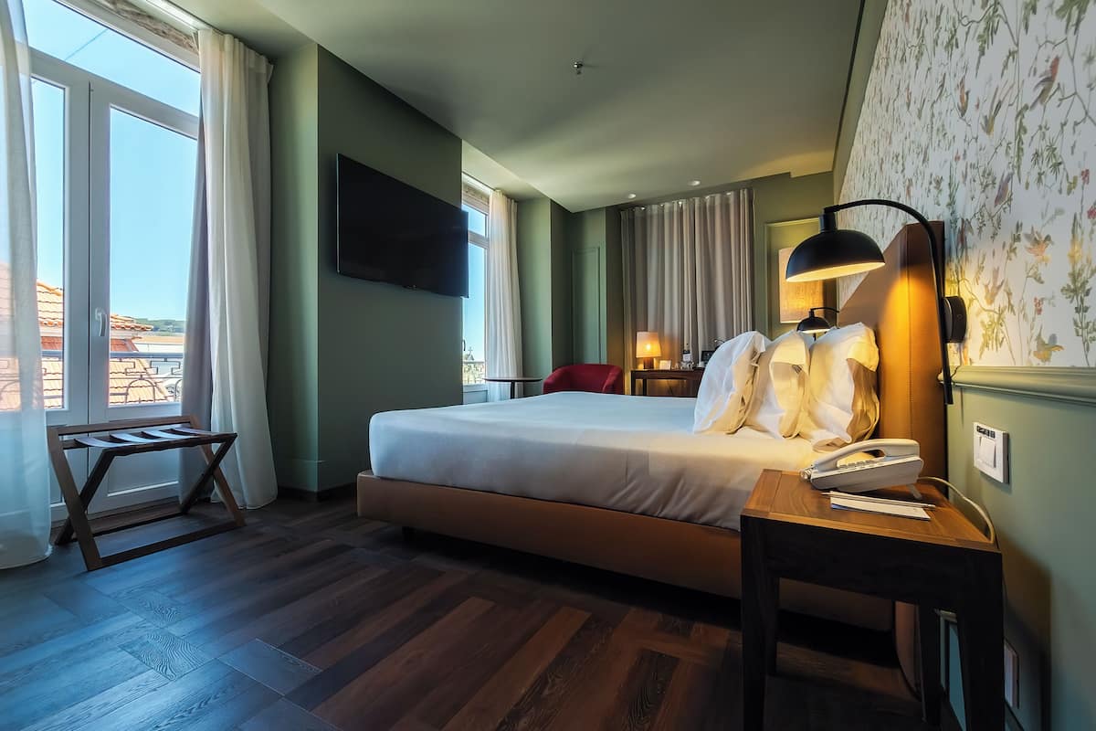 Quarto de hotel com cama, mesa de cabeceira, luminária e televisão fixada na parede.