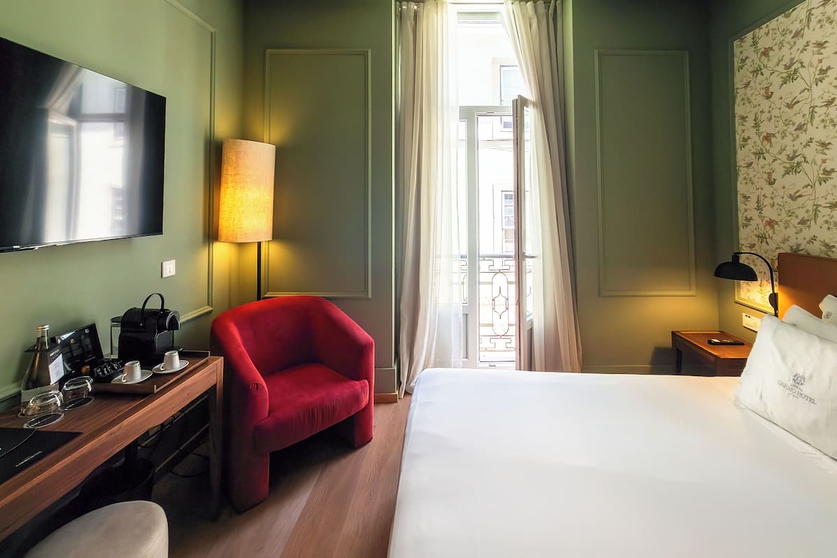Quarto de hotel com cama, escrivaninha com cadeira, televisão, abajur e janela com cortinas.