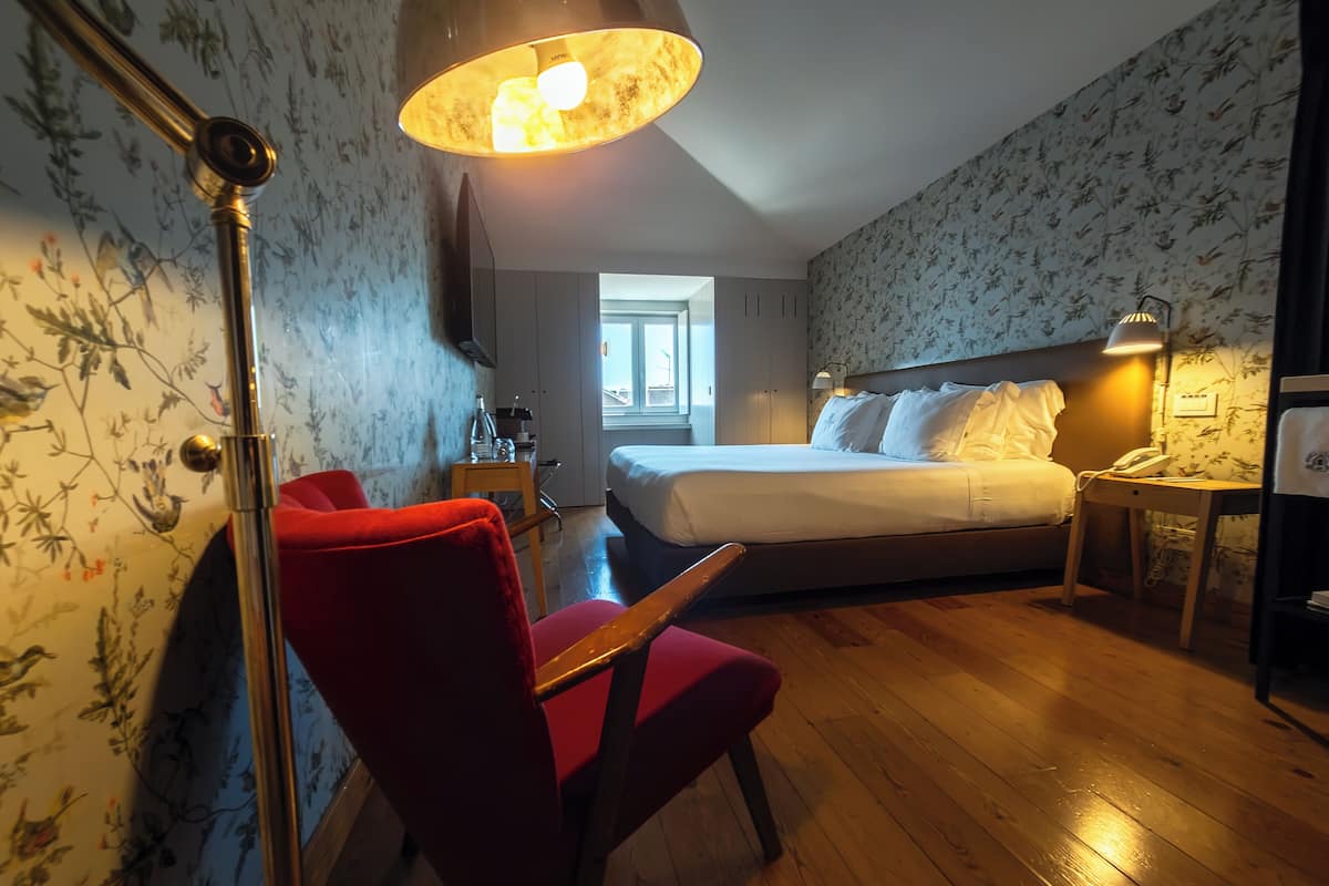 Quarto de hotel com cama, uma cadeira vermelha, escrivaninha com luminária e televisão na parede.