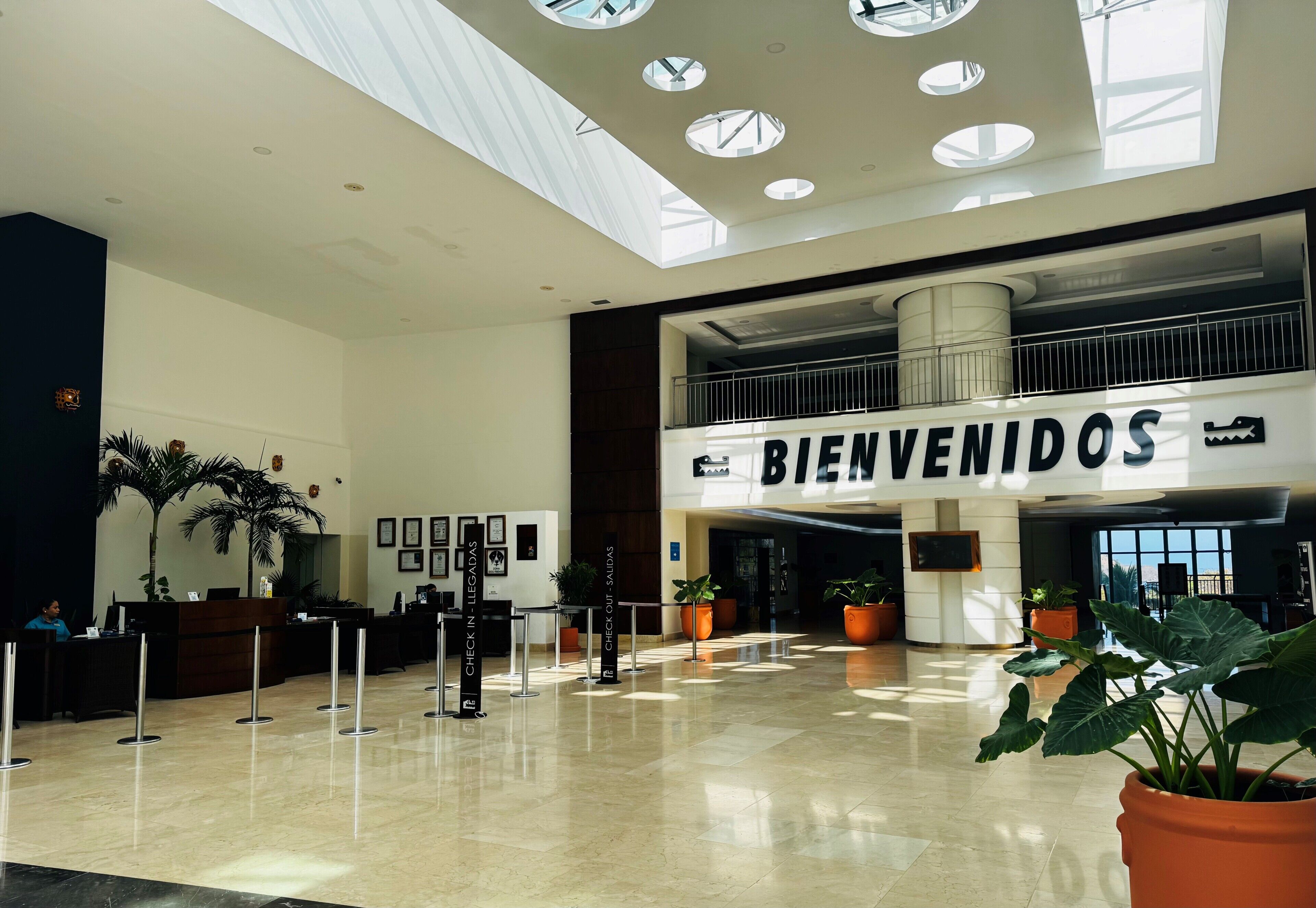 Um hall amplo com um letreiro 'BIENVENIDOS', balcão de recepção e plantas em vasos.