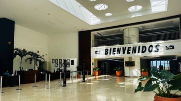 Um hall amplo com um letreiro 'BIENVENIDOS', balcão de recepção e plantas em vasos.