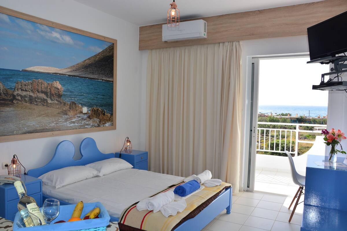 Quarto com cama, mesa de cabeceira, um aparador com garrafa e copos, pintura de uma paisagem costeira e varanda com vista para o mar.