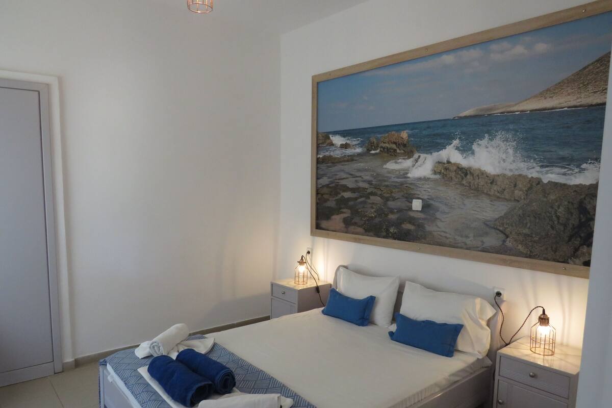 Quarto com cama, duas mesas de cabeceira, um quadro de uma praia e uma porta.