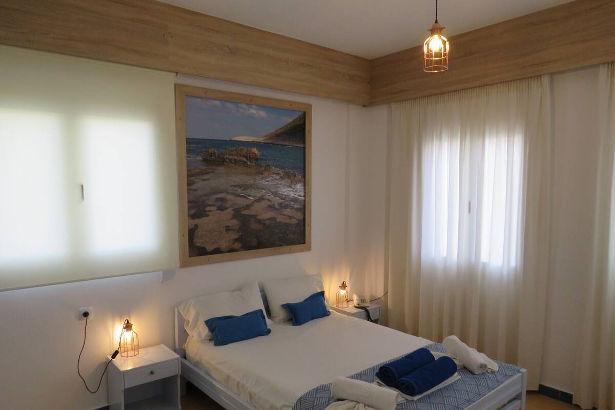 Quarto bem arrumado com cama, mesas de cabeceira, um quadro e uma janela com cortinas.