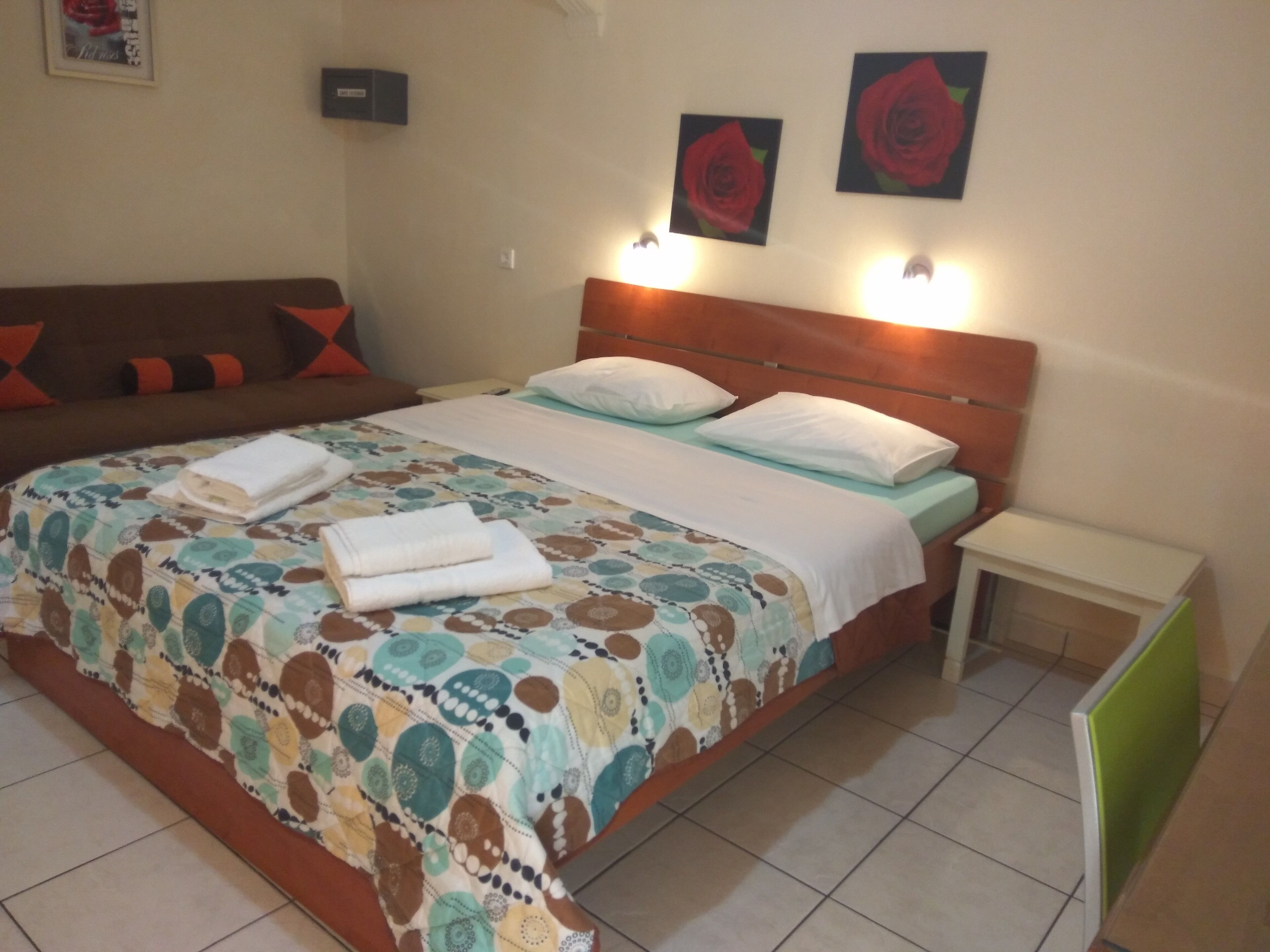 Cama bem arrumada com colcha colorida, cabeceira de madeira e duas luminárias fixadas na parede.