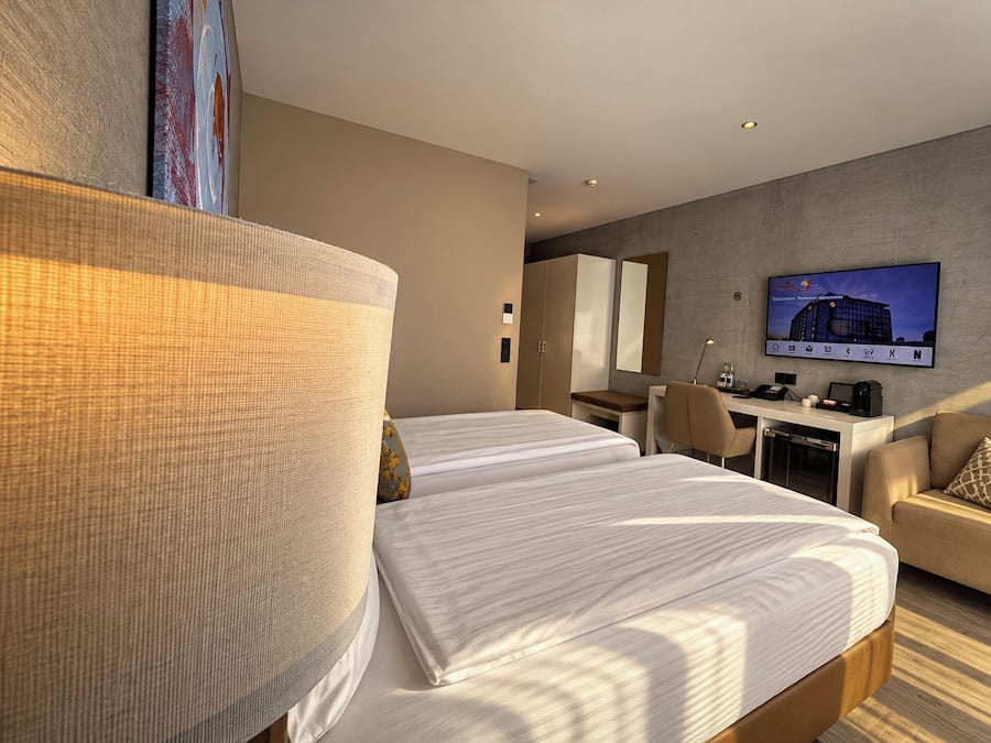 Quarto de hotel com duas camas, uma escrivaninha, uma cadeira, uma televisão e um quadro grande na parede.
