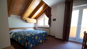Quarto com teto de madeira, cama com colcha floral, janela com cortinas e radiador.