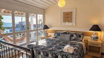Quarto com uma cama grande, duas luminárias de cabeceira, uma obra de arte emoldurada na parede e vista para o mar através de grandes janelas.