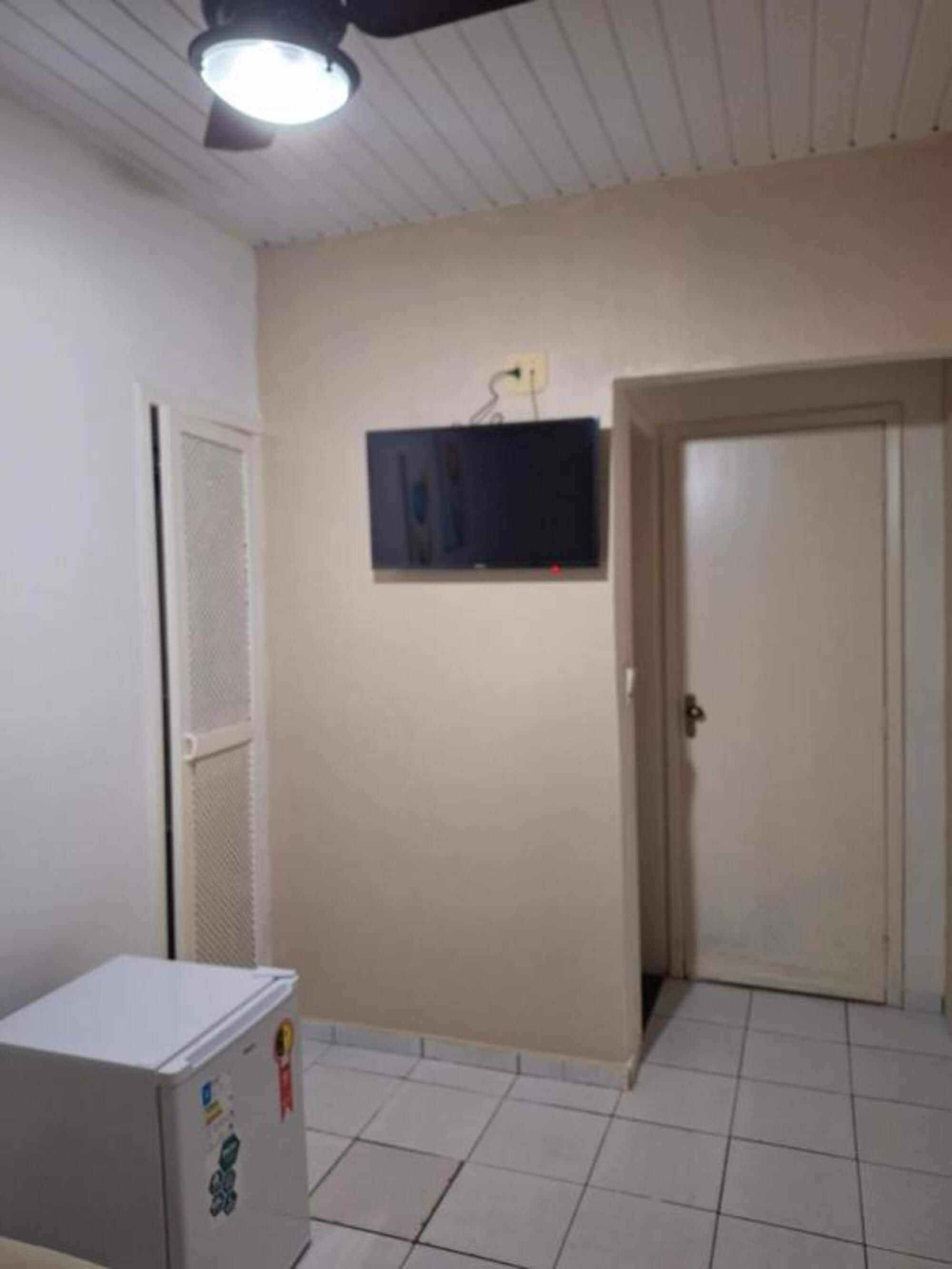 Quarto com chão em azulejos, porta, televisão na parede e pequeno frigorífico.