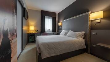 Quarto de hotel com uma cama grande, uma escrivaninha com cadeira, televisão e duas luminárias de parede.