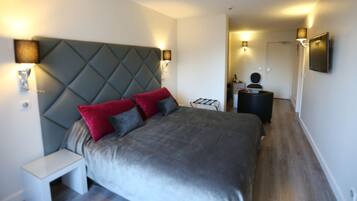 Quarto moderno com uma cama grande, mesa de cabeceira, cadeira e escrivaninha. Há luminárias fixadas na parede e uma TV de tela plana na parede.