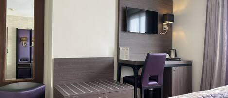 Quarto de hotel com cama, escrivaninha com cadeira, televisão e luminária fixada na parede.