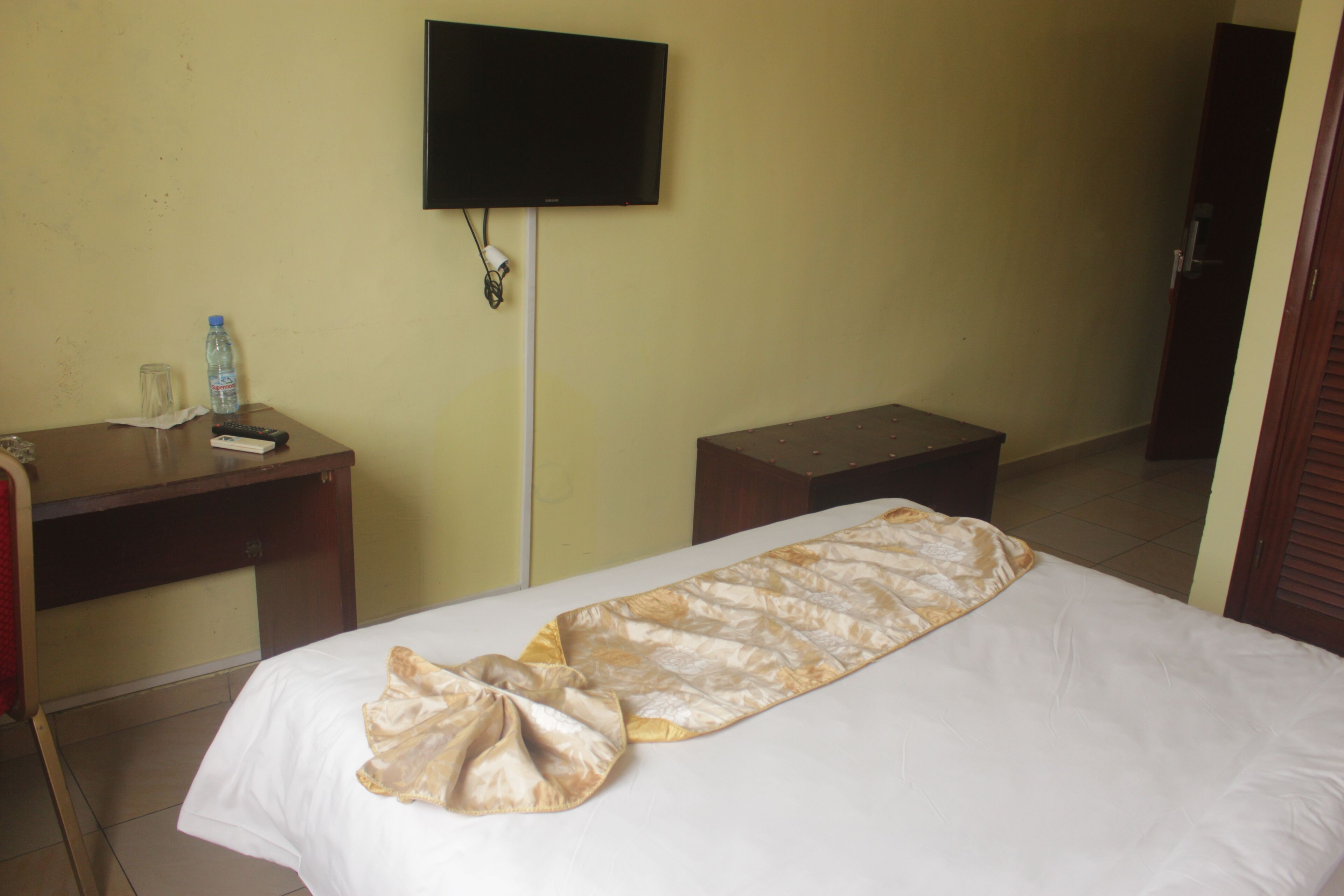 Quarto de hotel com cama, escrivaninha, televisão fixada na parede e uma garrafa de água sobre a escrivaninha.
