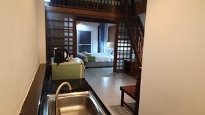 Uma cozinha estreita com uma pia, um eletrodoméstico verde e uma cadeira de madeira. Uma cama com travesseiro verde é visível através de uma porta no fundo.