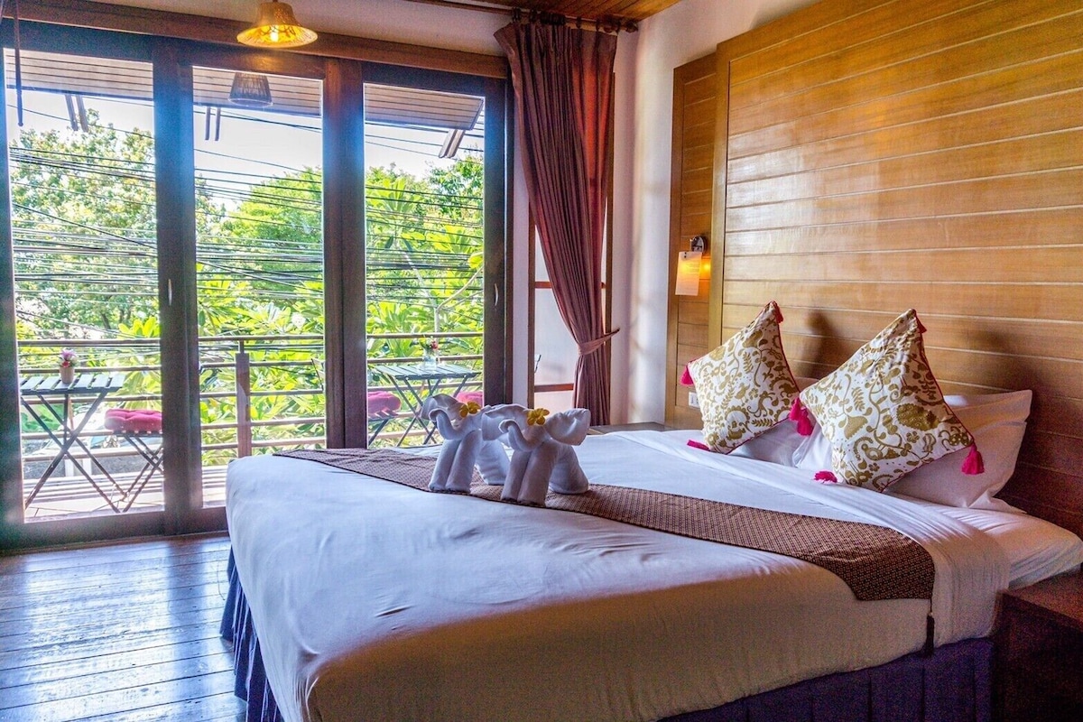 Quarto de hotel com cama, duas almofadas, mesa de cabeceira, janela com cortinas e vista para o verde lá fora.