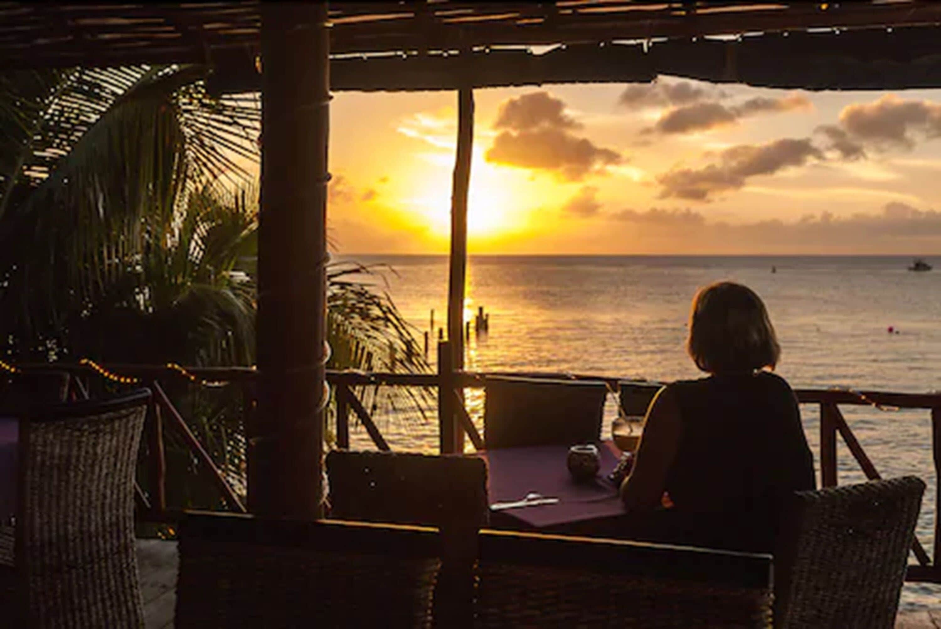 Um restaurante à beira-mar com mobiliário de vime e vista para o pôr do sol sobre o oceano.