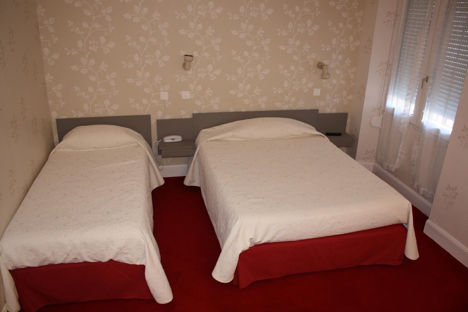 Quarto com duas camas de solteiro, tapete vermelho, papel de parede floral e janela com persianas.