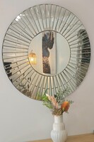 Espelho redondo com design radial numa parede, refletindo uma mão e um candeeiro.