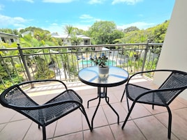Uma varanda com duas cadeiras de vime preto, uma mesa redonda pequena com uma planta em vaso, e vista para uma piscina e vegetação.