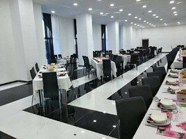 Uma ampla sala de conferências com longas mesas preparadas para refeição, equipada com cadeiras e mesas, e um teto moderno com iluminação embutida.