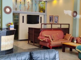 Um lobby com uma mesa de recepção, uma televisão fixada na parede, um sofá, uma poltrona vermelha e uma mesa de centro.