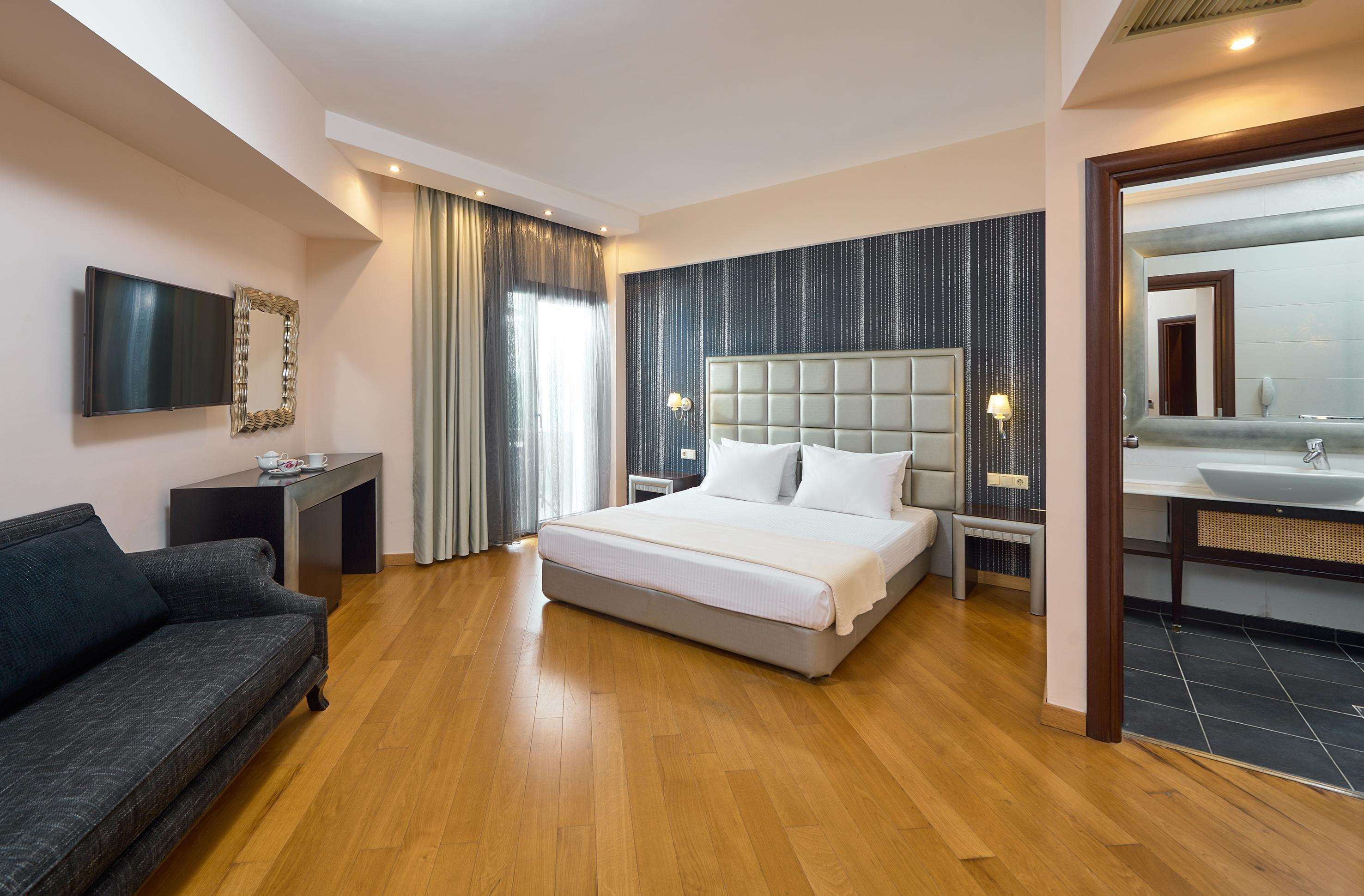 Quarto de hotel moderno com uma cama grande, um sofá, uma TV e um banheiro com um espelho amplo.