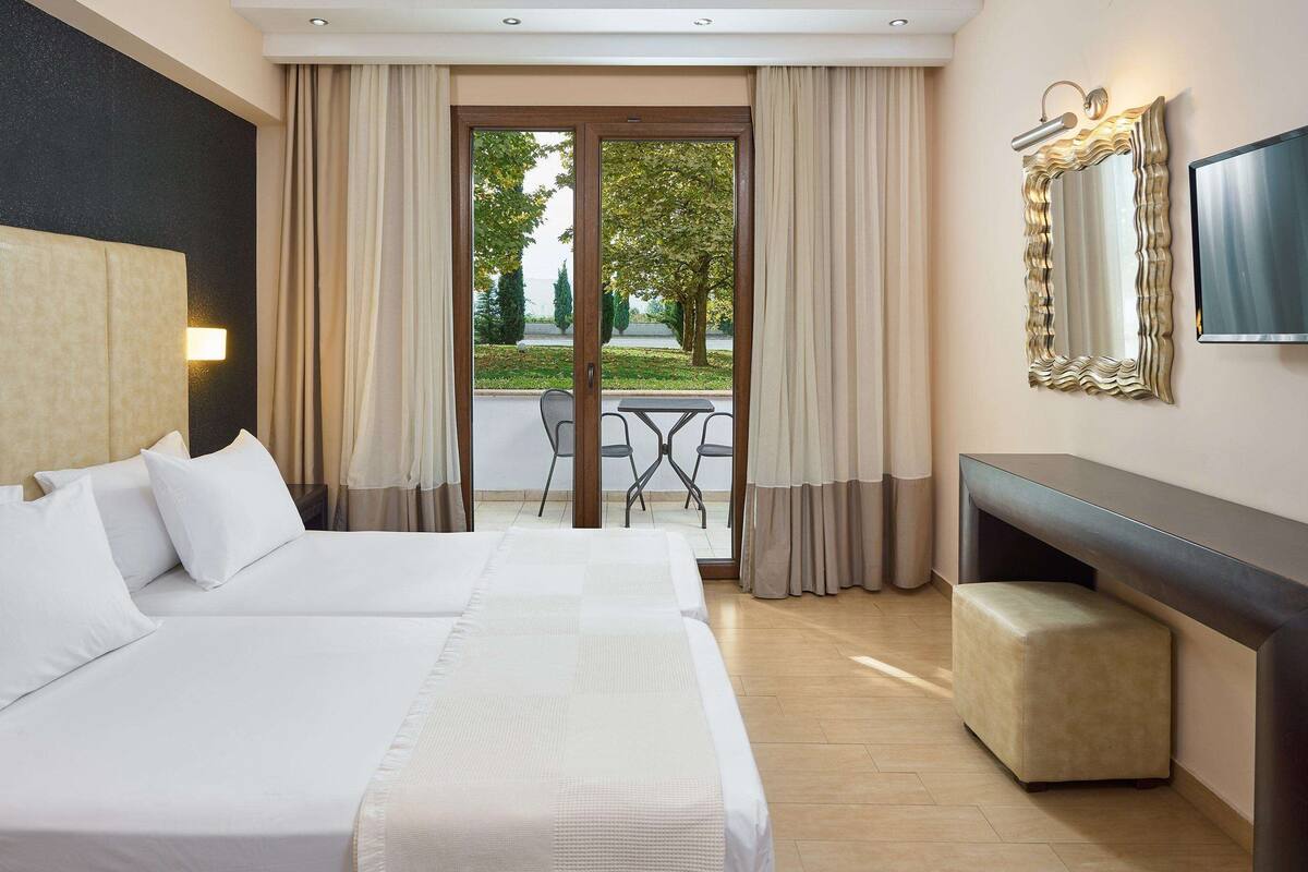 Quarto de hotel com duas camas, televisão, escrivaninha e vista para o exterior.