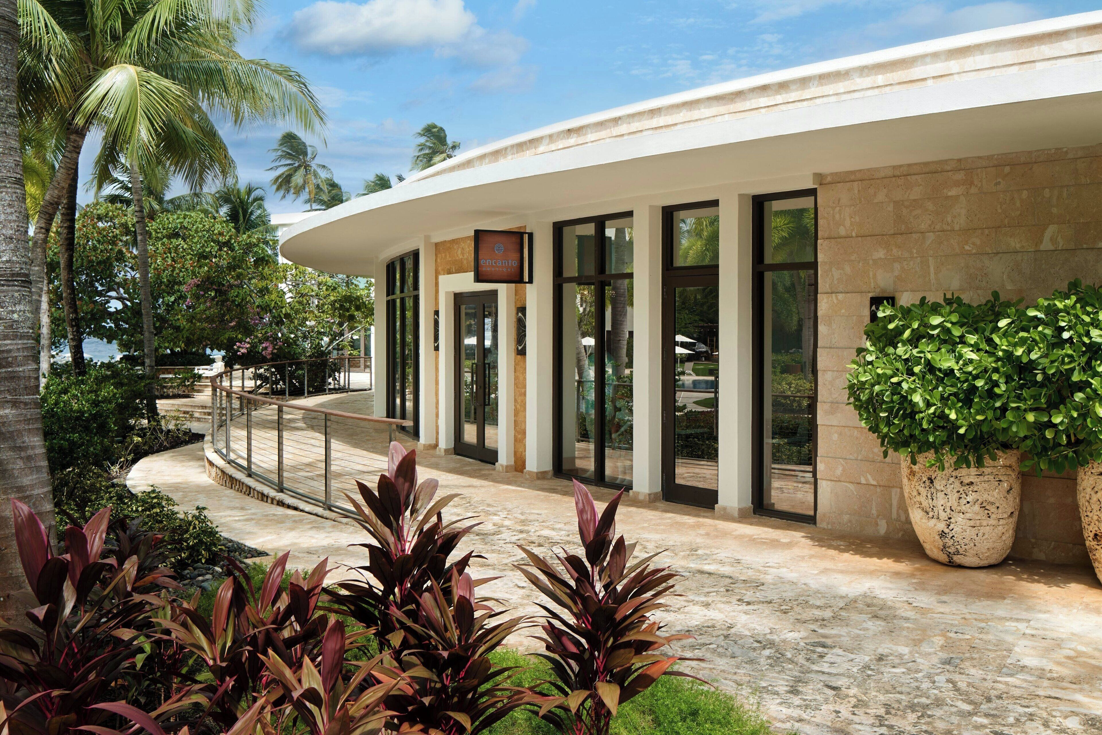 Entrada de hotel com galeria coberta, palmeiras e plantas em vasos.