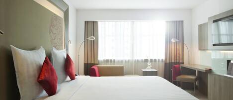 Quarto de hotel com uma cama grande, escrivaninha, cadeira, televisão e janela com cortinas.