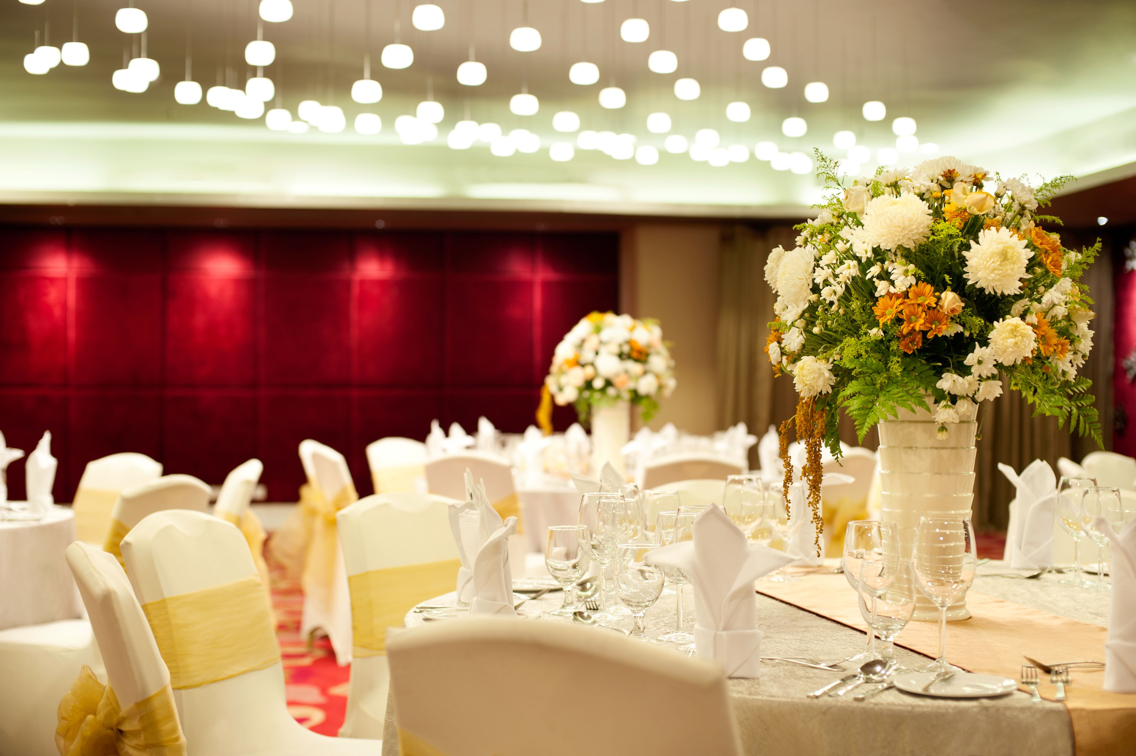 Uma sala de banquetes com mesas redondas preparadas para refeições, cadeiras brancas com fitas amarelas e arranjos florais.