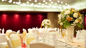 Uma sala de banquetes com mesas redondas preparadas para refeições, cadeiras brancas com fitas amarelas e arranjos florais.