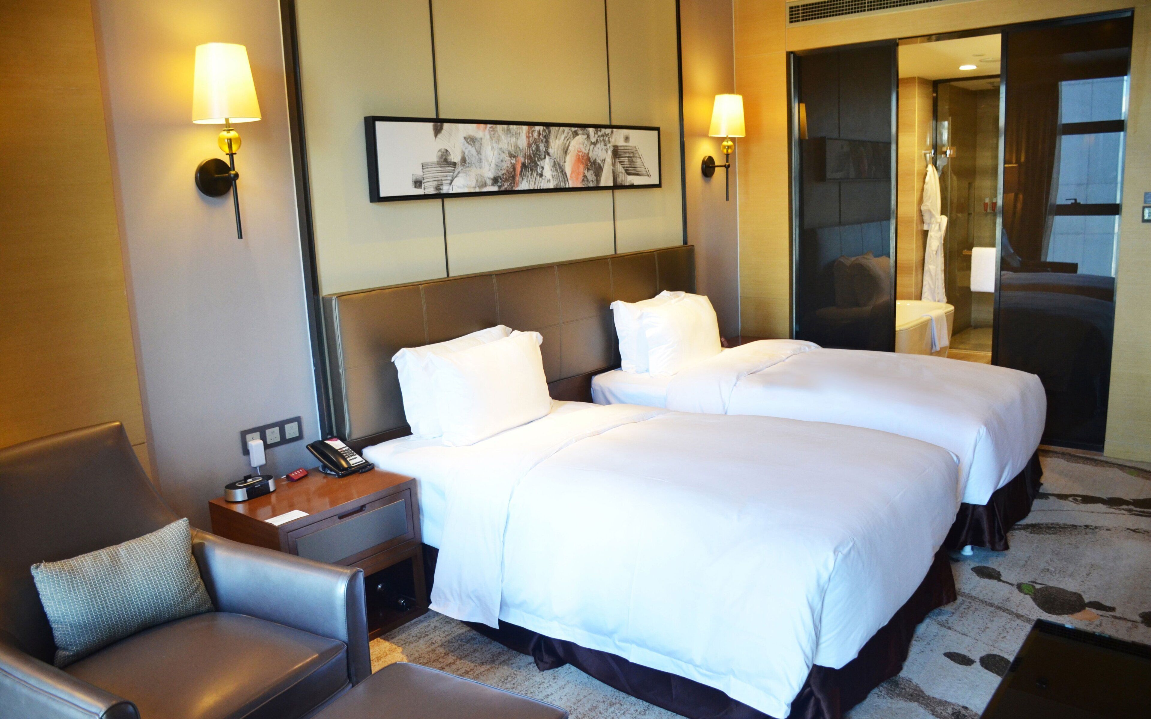 Quarto de hotel com duas camas, uma escrivaninha com telefone, uma cadeira e um quadro na parede.