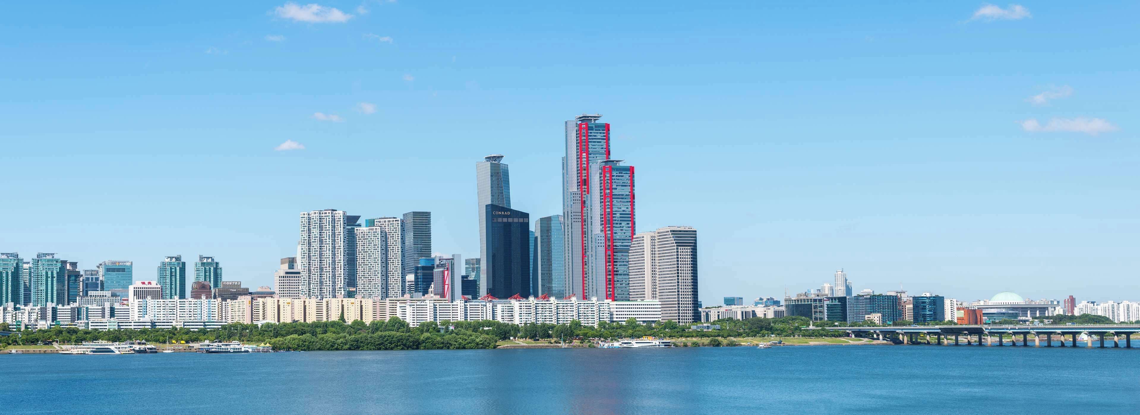 Horizonte urbano com edifícios modernos altos, um rio em primeiro plano e uma ponte à distância.